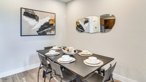 Uma sala de jantar com uma mesa retangular posta para seis pessoas, rodeada por seis cadeiras. Há duas obras de arte na parede e um espelho.