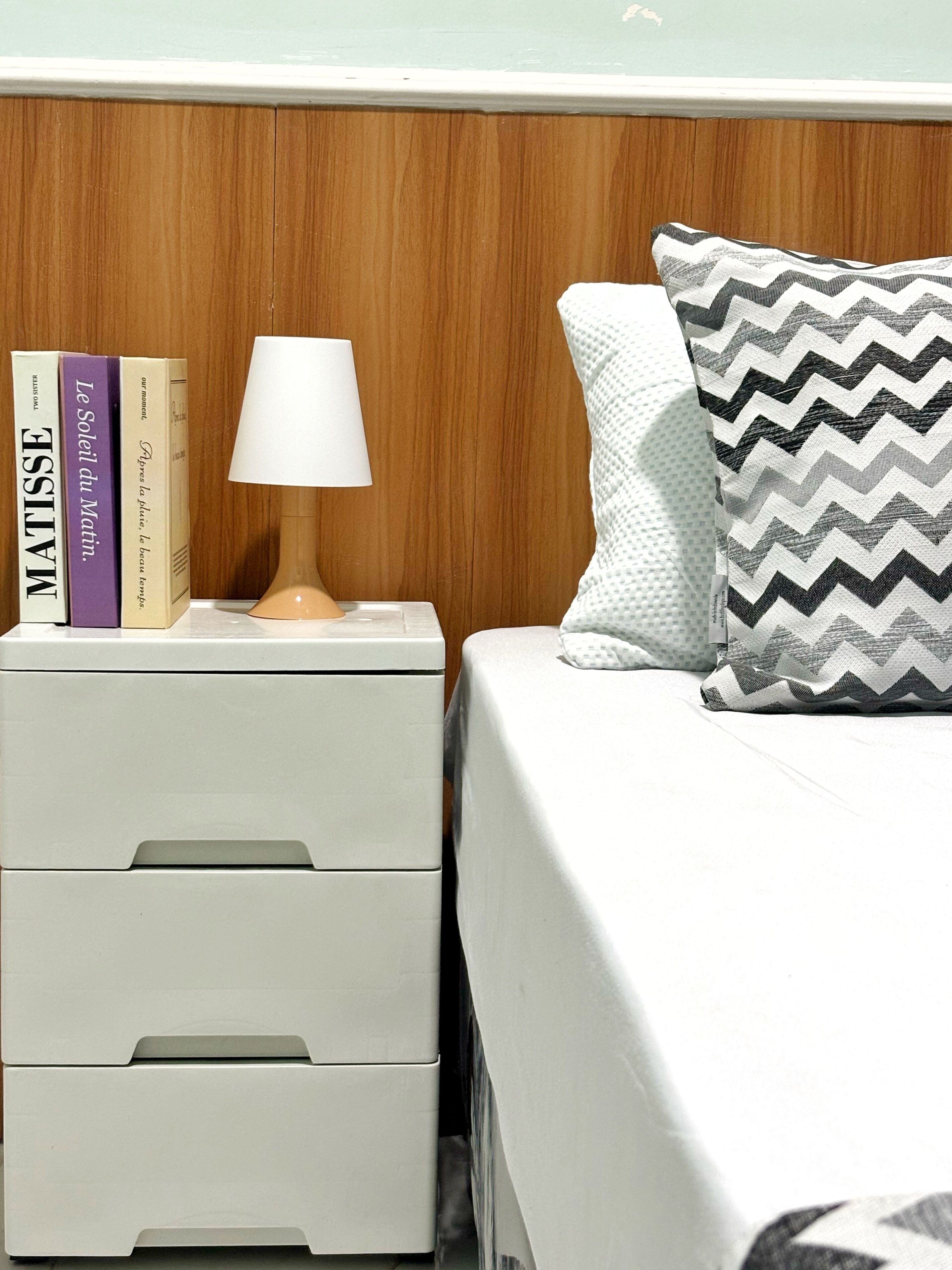 Cama bem arrumada com travesseiro cinza e branco em padrão ziguezague, abajur e pilha de livros, incluindo 'Matisse', sobre um criado-mudo branco encostado em cabeceira de madeira.