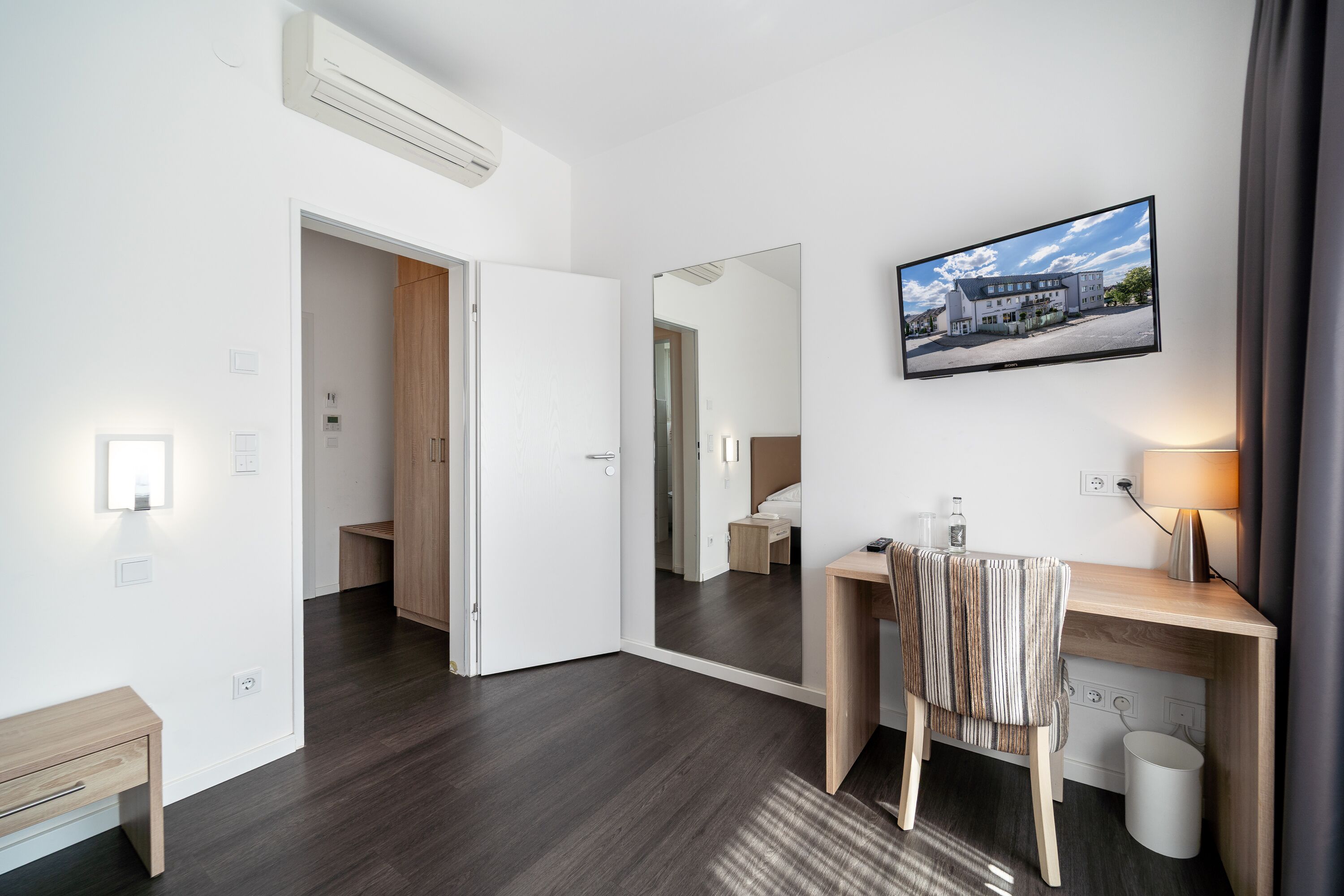 Quarto de hotel moderno com escrivaninha de madeira, uma cadeira, um espelho, televisão e uma mesinha de cabeceira.