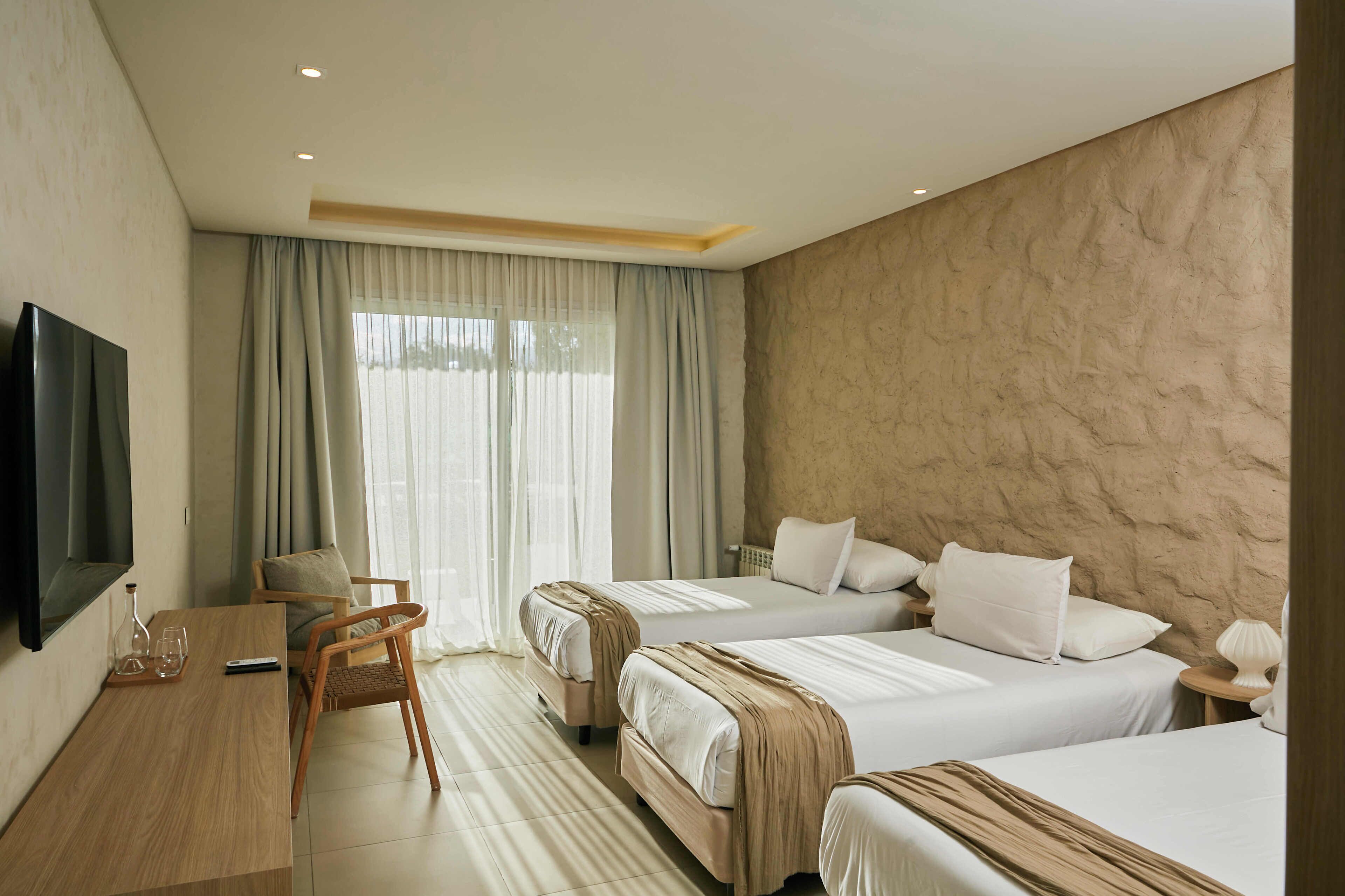 Quarto de hotel com duas camas, uma escrivaninha, uma cadeira, uma televisão e uma janela grande com cortinas.