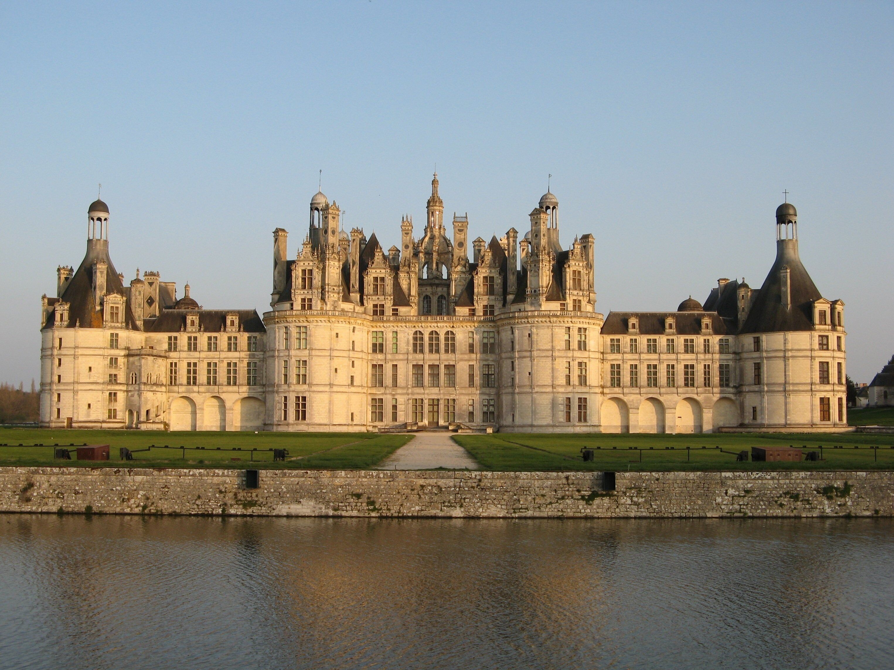 Um edifício grande e histórico, com várias torres e fachada simétrica, localizado próximo a um corpo d'água.