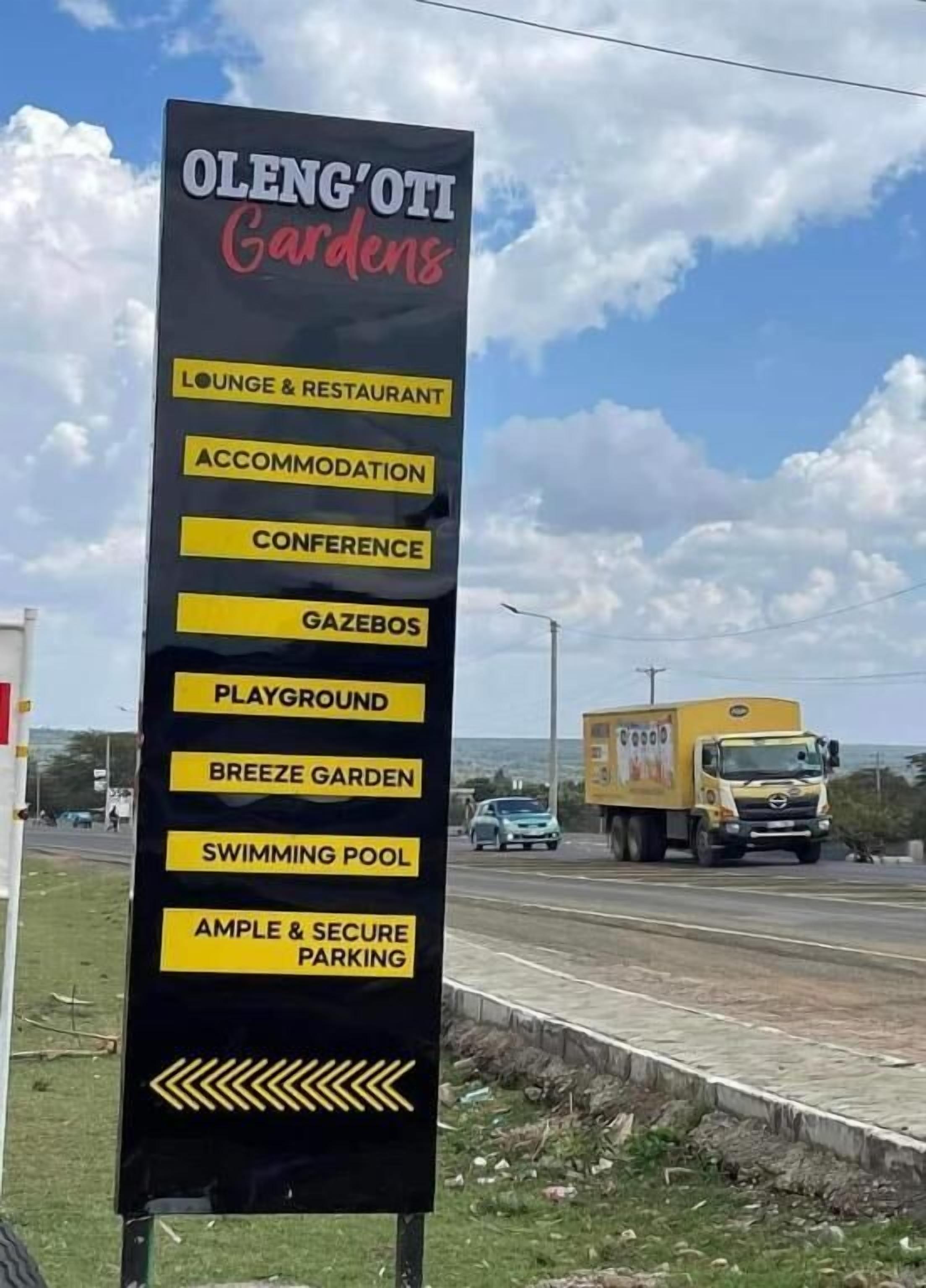 Placa de Oleng'oti Gardens listando comodidades como lounge, restaurante, acomodações, instalações para conferências, gazebos, parquinho, jardim com brisa, piscina e amplo estacionamento seguro.