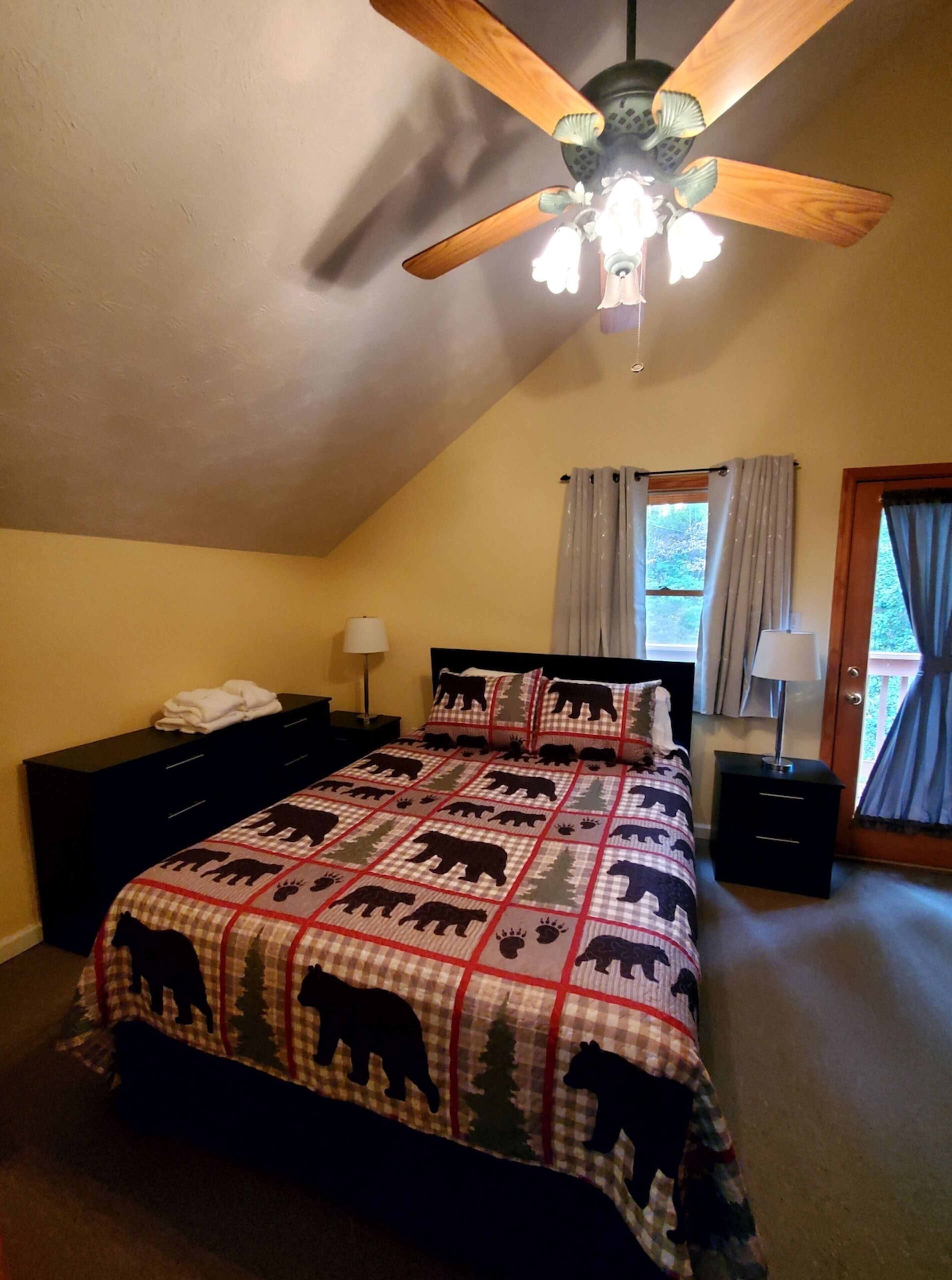 Quarto com teto inclinado, cama com colcha temática de urso, ventilador de teto e mesa de cabeceira com abajur.