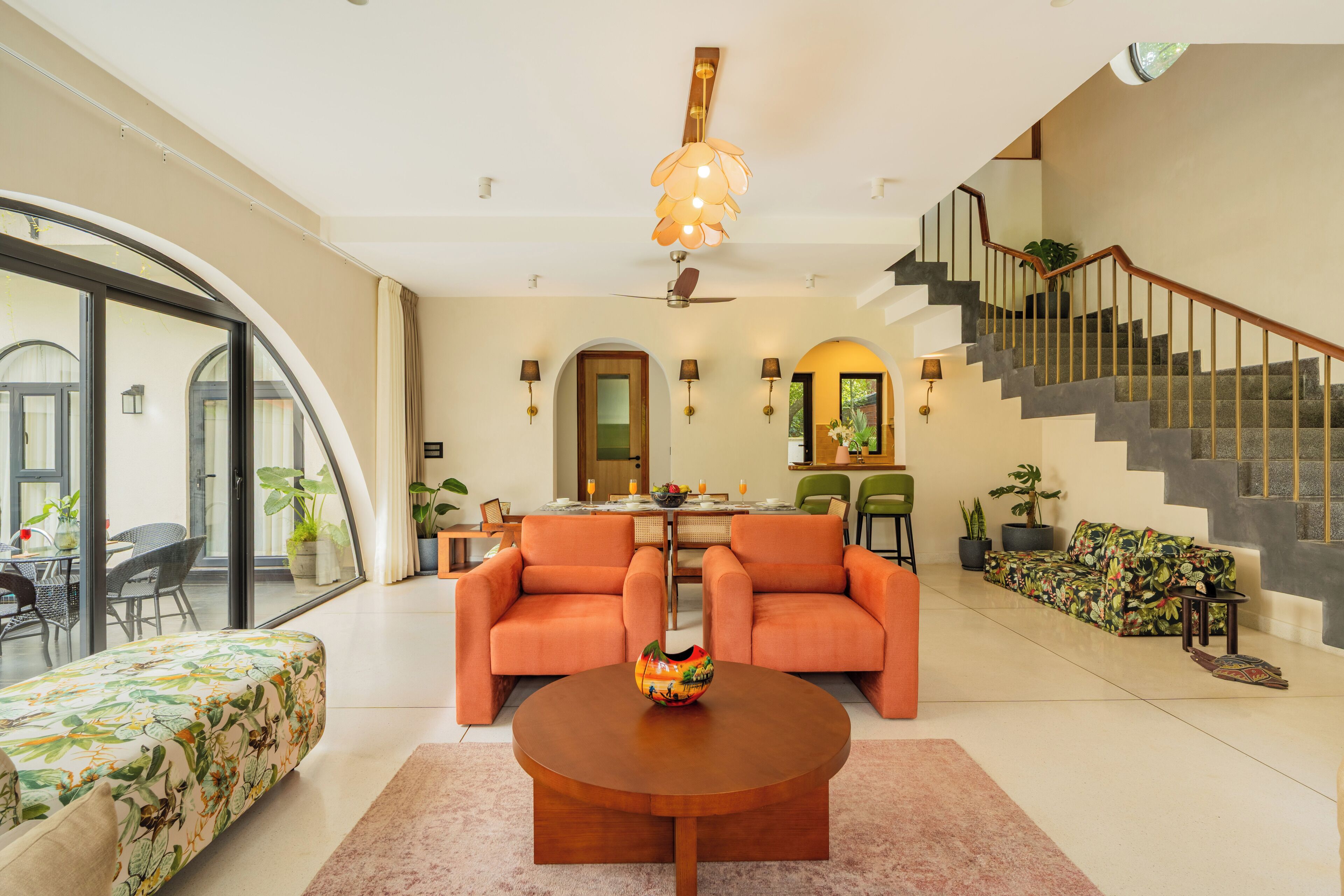 Uma sala de estar ampla com uma mesa de centro redonda de madeira, duas poltronas laranja, uma mesa de jantar com cadeiras e uma escada com corrimão.