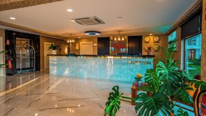 Uma área de recepção de hotel com balcão de mármore, relógios e plantas em vasos.