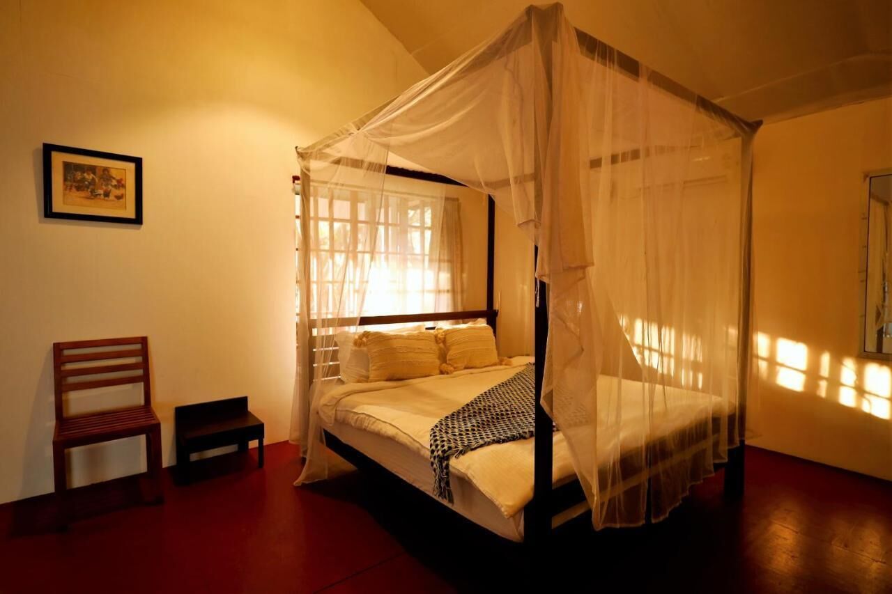 Quarto com cama de dossel, uma cadeira de madeira, um pequeno banco e um quadro pendurado na parede.
