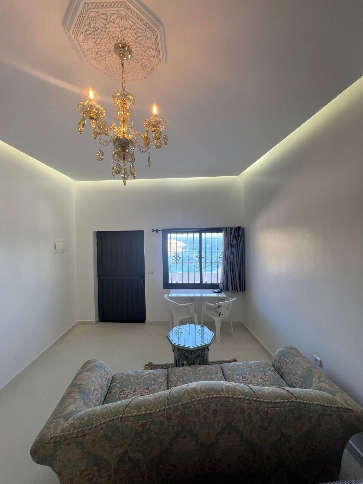 Uma sala de estar com um candelabro, um sofá, uma mesa pequena e uma janela com cortinas.