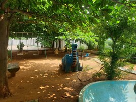 Um parquinho infantil com um escorregador azul e balanços, cercado por árvores e um chão de terra.