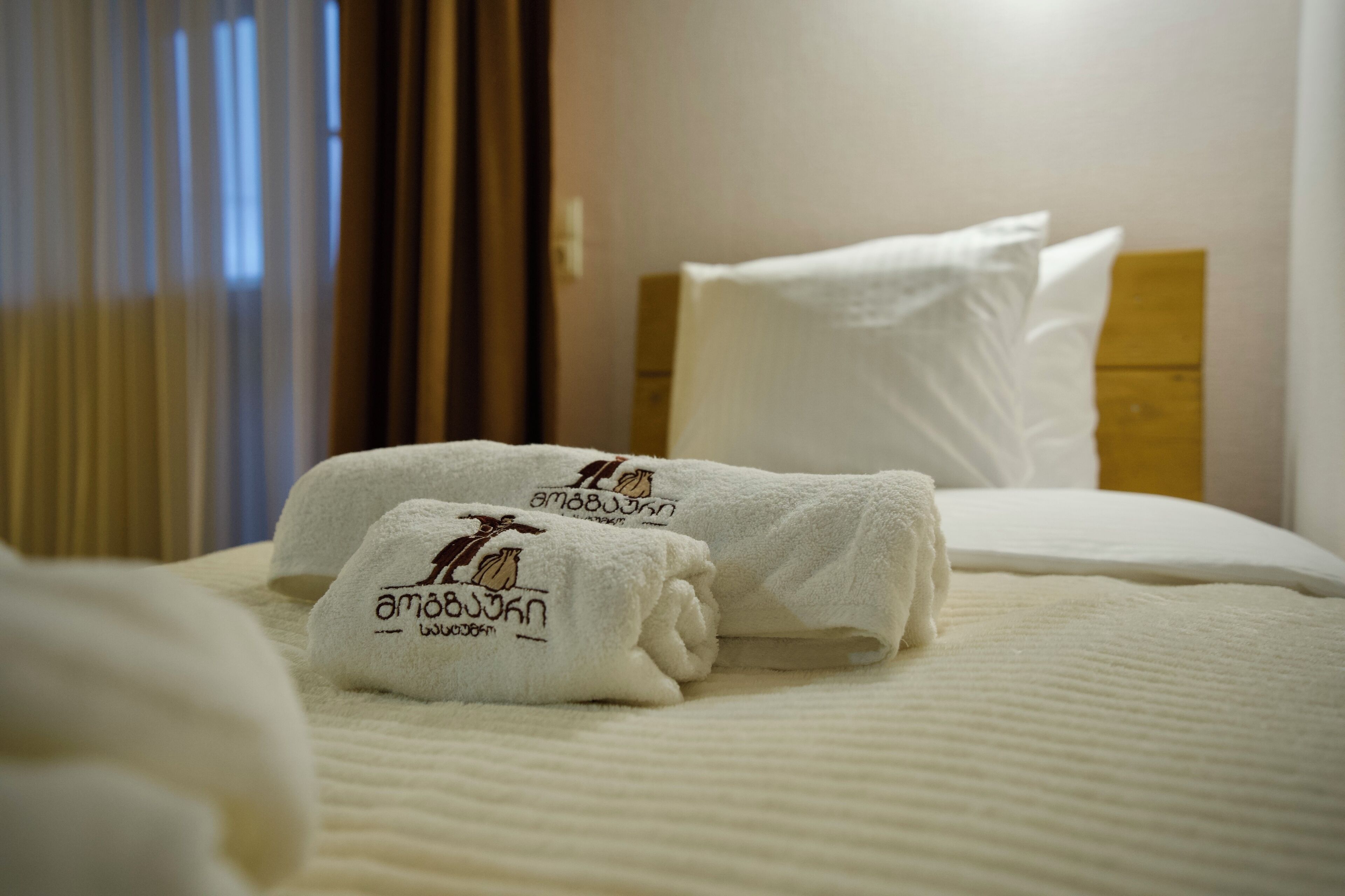 Cama bem arrumada com duas toalhas com logotipo e texto, cabeceira de madeira e janela com persianas.