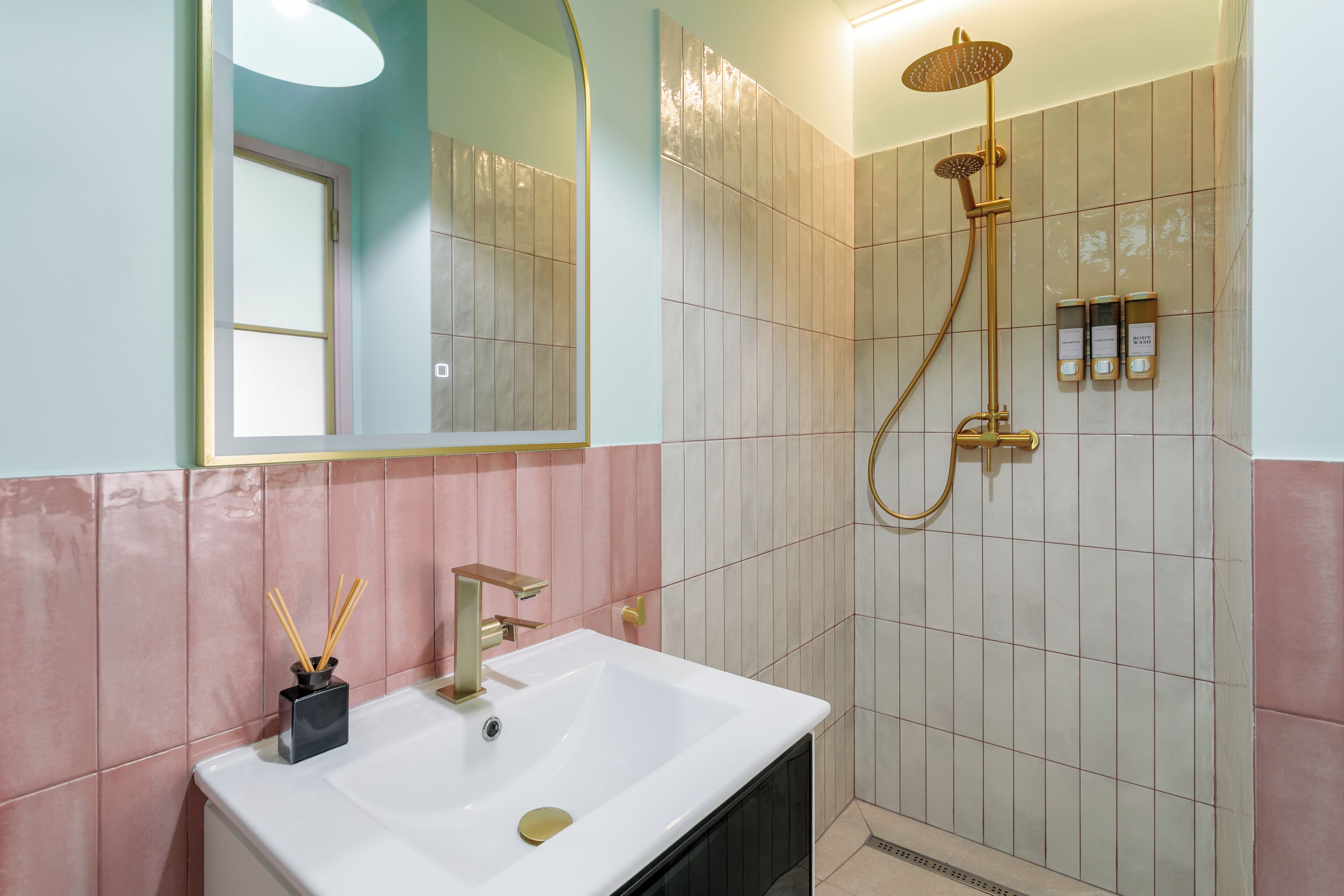 Casa de banho com lavatório branco, espelho, chuveiro dourado e paredes revestidas a azulejos rosa e brancos.