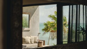 Vista emoldurada de um quarto com janela para a praia, um sofá e uma mesinha.
