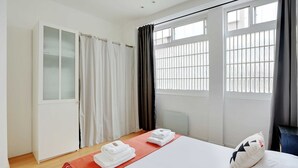 Quarto de hotel com cama, manta vermelha, travesseiro branco e janela com persianas.
