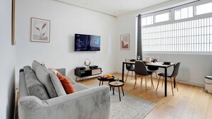 Uma sala de estar moderna com um sofá cinza, uma mesa de jantar com cadeiras, uma TV fixada na parede e uma grande janela com persianas.