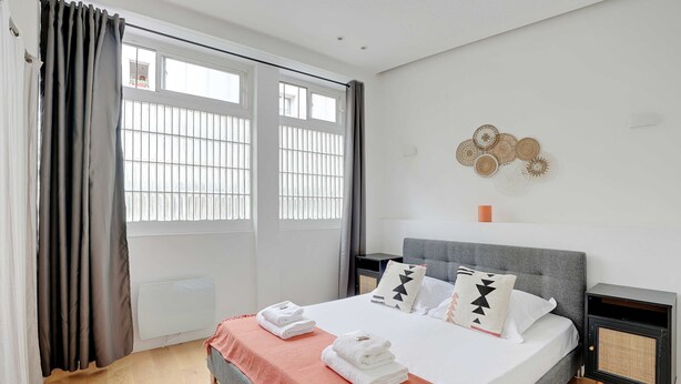 Quarto de hotel moderno com uma cama grande, cabeceira cinza e mesa de cabeceira com cesto trançado.
