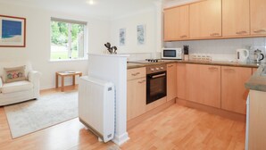 Uma cozinha moderna com armários de madeira, forno embutido, micro-ondas e chaleira. Há um radiador branco, uma mesa pequena e um sofá branco na sala de estar.