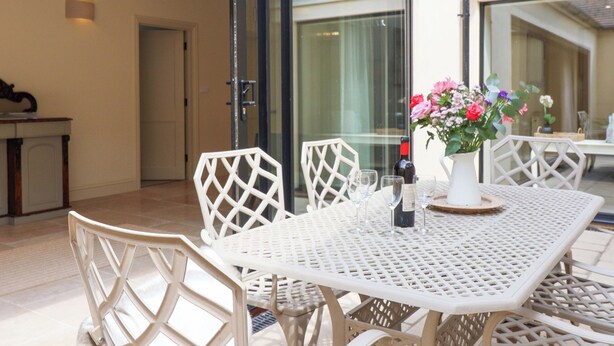 Um pátio externo com mesa e cadeiras brancas, um vaso de flores e uma garrafa de vinho.