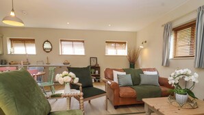Uma sala de estar aconchegante com uma poltrona verde, um sofá de couro marrom e uma mesa de centro de madeira. Há um vaso com flores na mesa e uma janela com persianas ao fundo.