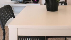 Um quarto com uma mesa branca, uma cadeira preta, um vaso com planta, um quadro de uma praia e uma televisão sobre um suporte.