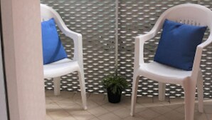 Uma varanda com duas cadeiras de plástico branco, cada uma com uma almofada azul, e um vaso de planta.