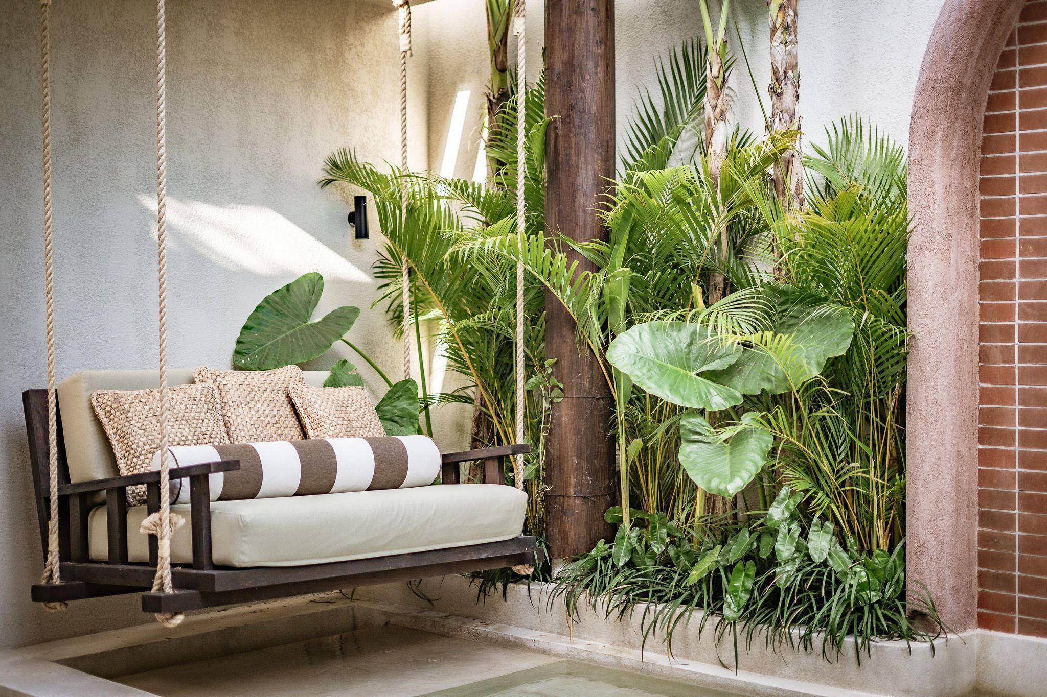 Uma cadeira balanço suspensa com almofada e travesseiros, cercada por plantas tropicais exuberantes e uma parede de tijolos.
