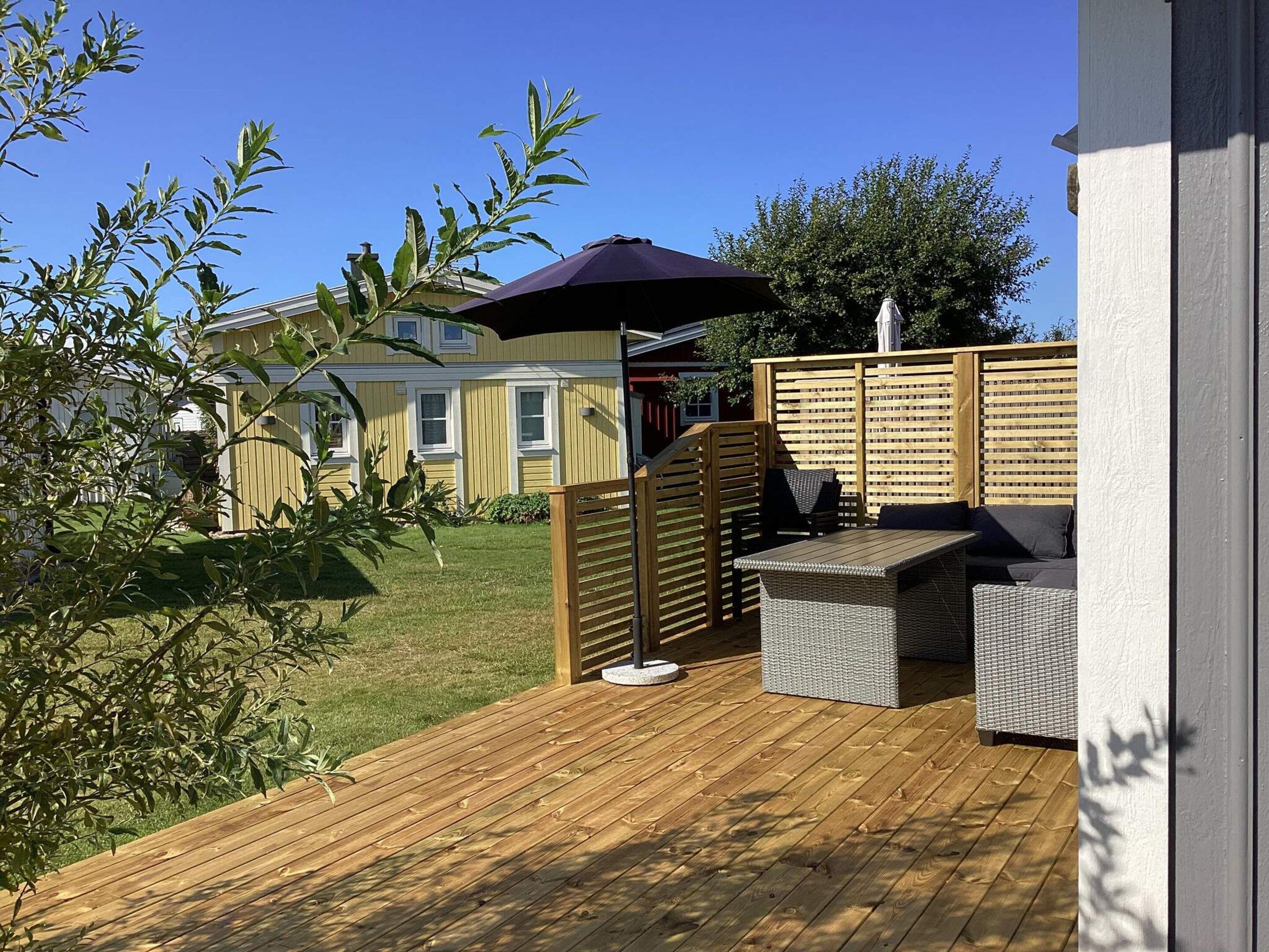 Um deck de madeira com móveis externos, uma pérgula e vista para uma casa.