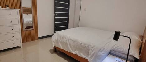 Quarto com cama, mesa de cabeceira, luminária, cômoda, espelho e porta com puxador vertical.