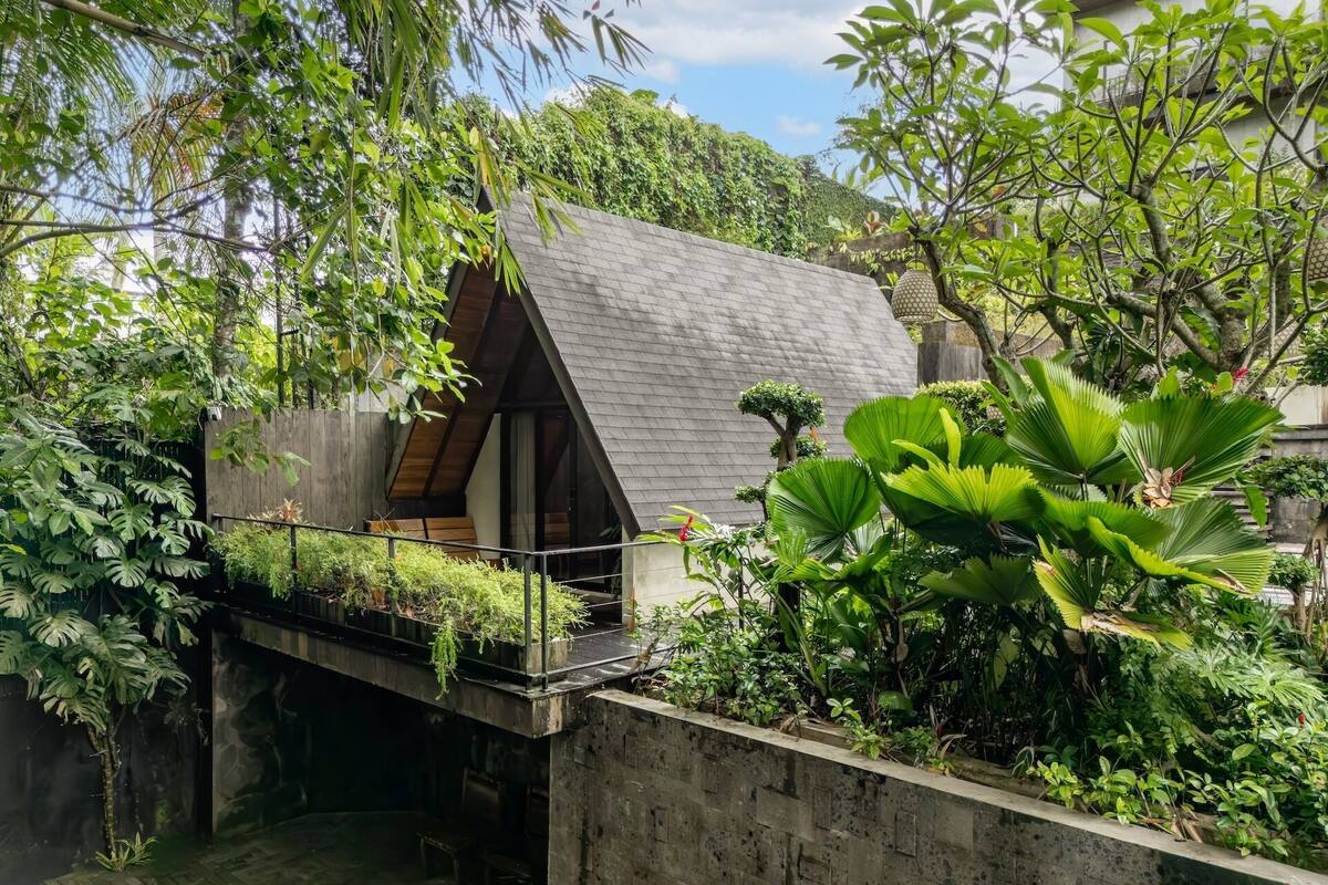 Uma casa moderna com telhado inclinado, rodeada por vegetação exuberante e um pequeno elemento aquático.