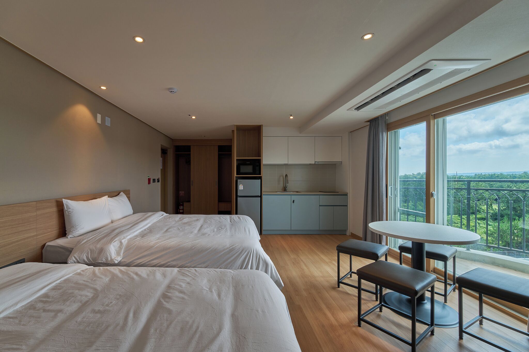 Quarto de hotel com duas camas, uma cozinha compacta, área de refeições com mesa e bancos, e varanda com vista.