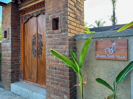 Uma porta de madeira com esculturas detalhadas, uma parede de tijolos e uma placa com a inscrição 'BUJANA SARI VILLA'.