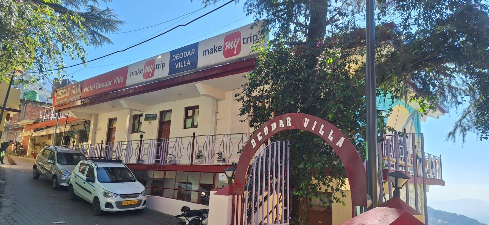 Entrada de hotel com uma entrada de automóveis gradeada, uma placa anunciando 'Hotel Deodar Villa' e serviços 'make my trip', e carros estacionados.