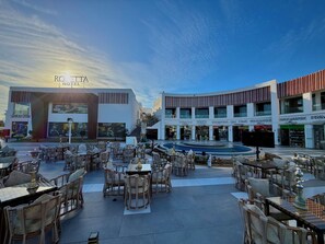 Uma área externa com mesas e cadeiras, piscina e entrada de hotel com sinalização.