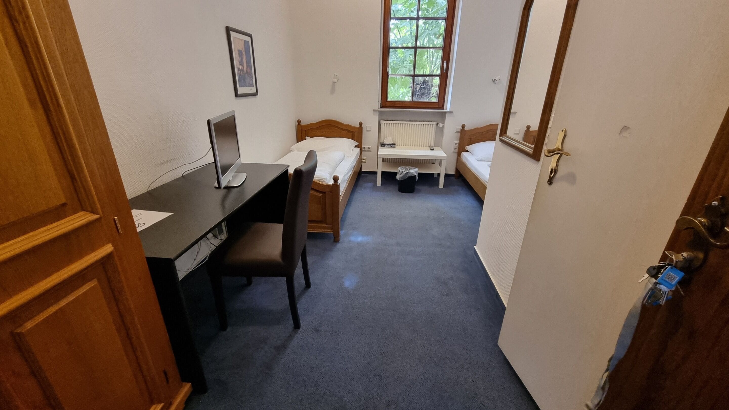 Quarto pequeno e funcional, com escrivaninha, duas camas de solteiro, janela com vista e porta com chave na fechadura.