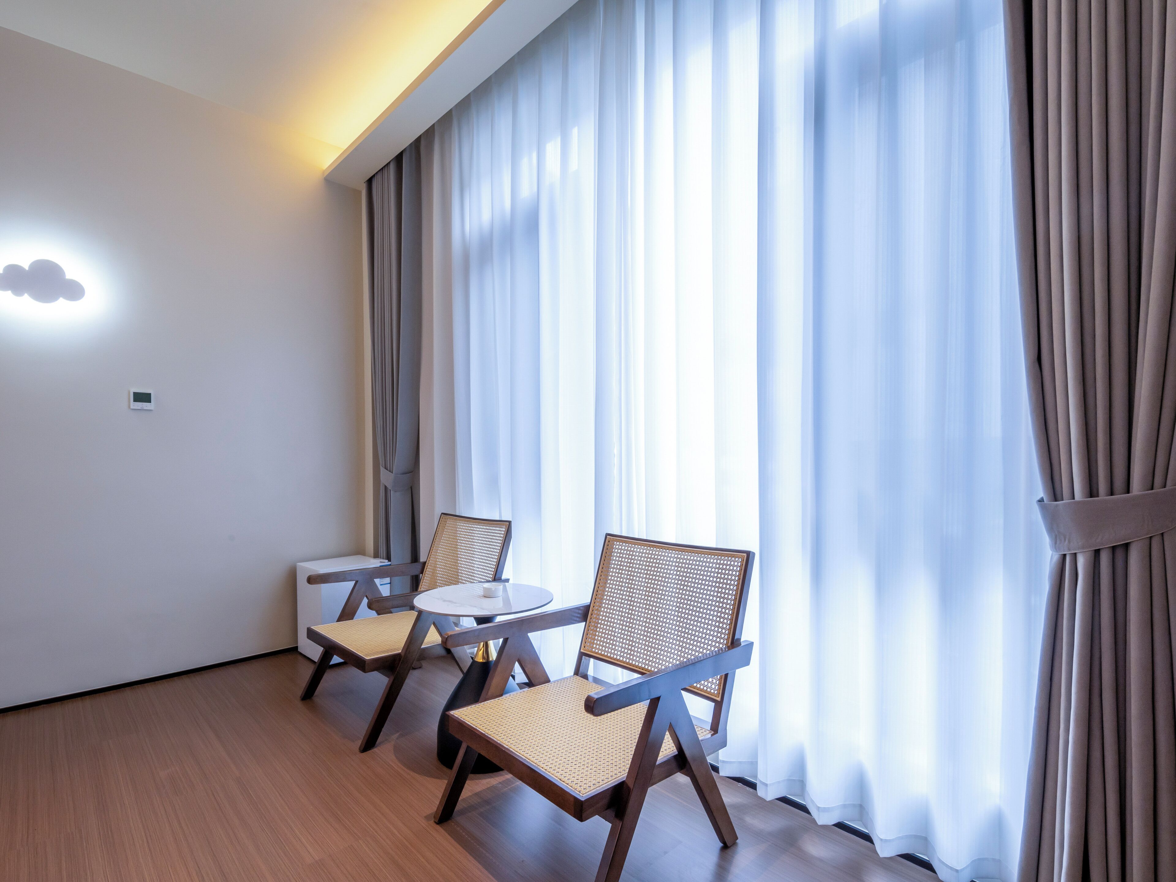 Quarto de hotel com duas cadeiras de madeira, uma mesa pequena e uma janela grande com cortinas finas.