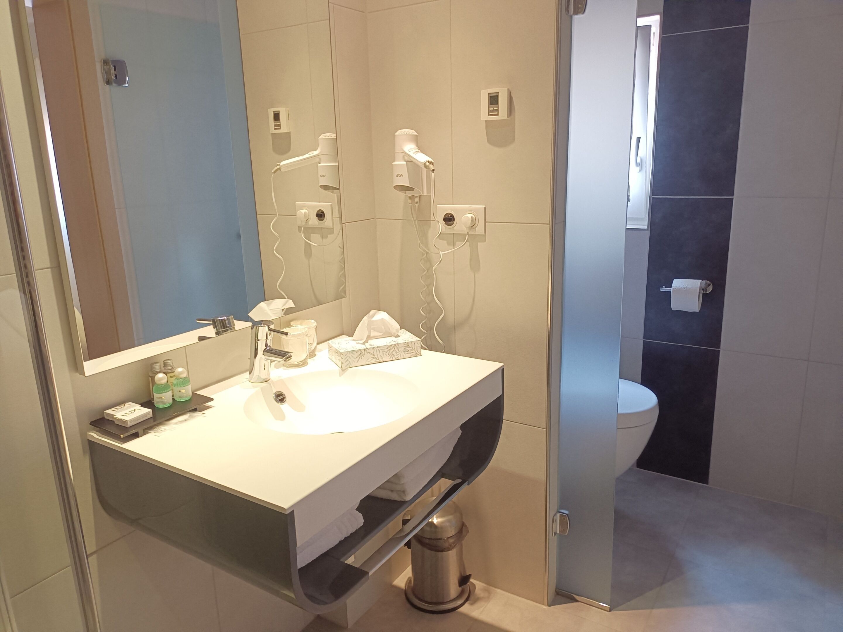 Um banheiro moderno com pia branca, dois secadores de cabelo, espelho e vaso sanitário.