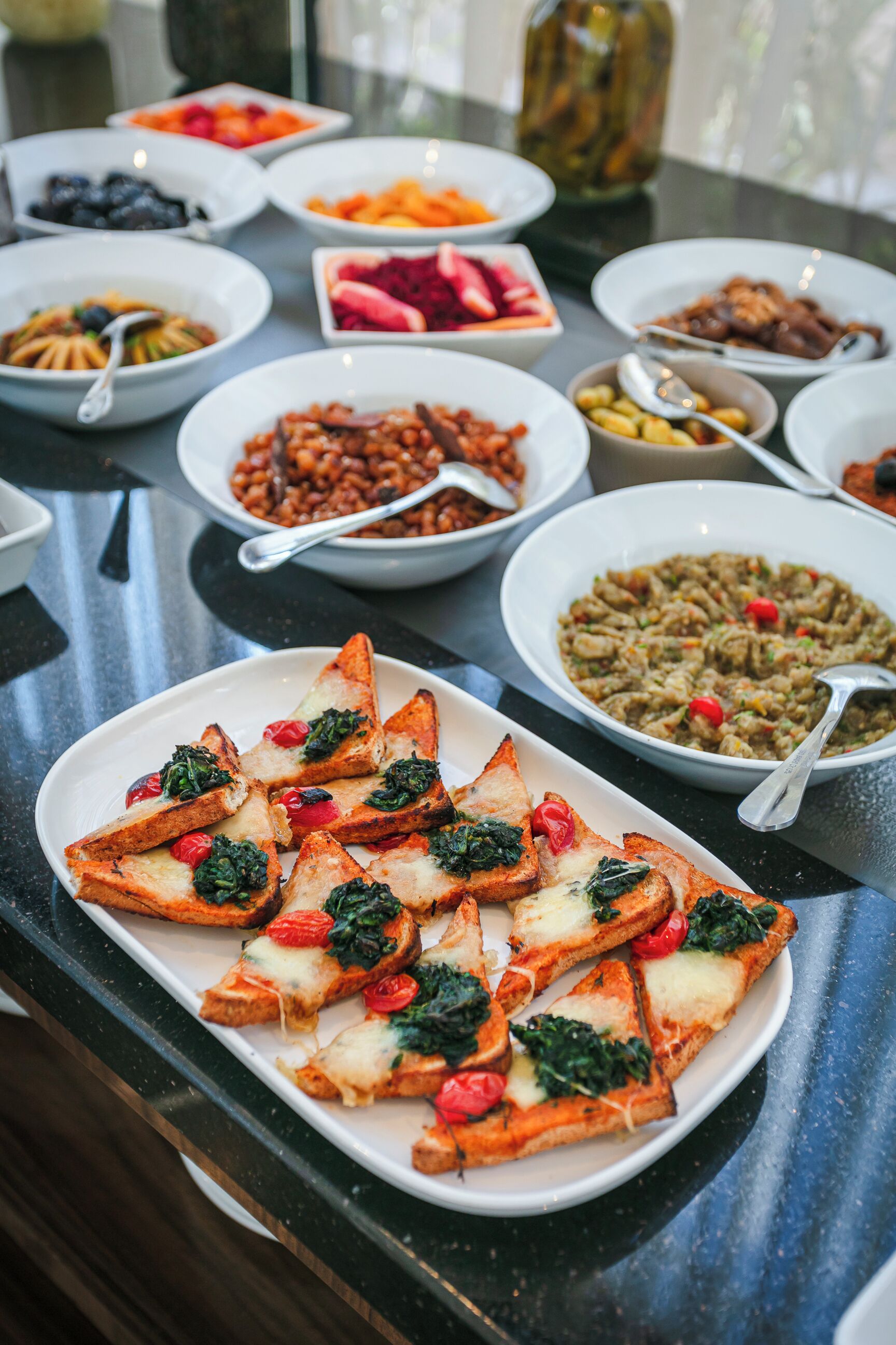 Mesa de buffet com diversos pratos, incluindo uma travessa de pão torrado com queijo e verduras, e várias tigelas com diferentes alimentos.