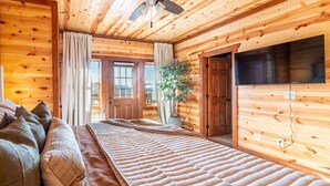 Quarto de cabana de madeira com cama, cortinas, ventoinha no teto e uma janela grande.