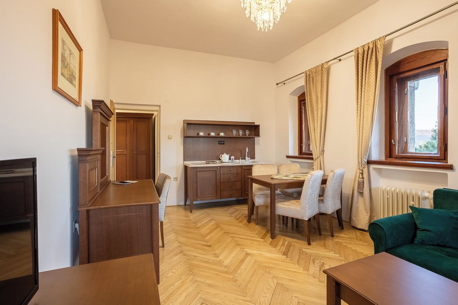 Um quarto com piso de madeira em padrão espinha de peixe, uma mesa de jantar com cadeiras, uma cozinha compacta, um sofá verde e uma janela com cortinas.