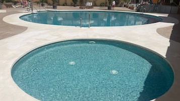 Uma grande piscina circular com borda em azulejos, rodeada por um espaço pavimentado com vasos de plantas e guarda-sóis.