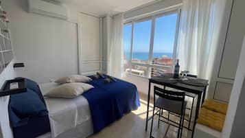 Quarto de hotel com uma cama, uma mesinha com uma garrafa e um copo, uma cadeira e vista para o mar e edifícios.