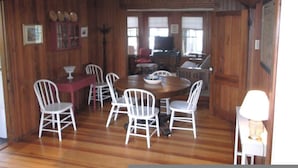 Uma sala de jantar com uma mesa de madeira e cadeiras brancas, uma televisão ao fundo, e painéis de madeira nas paredes.