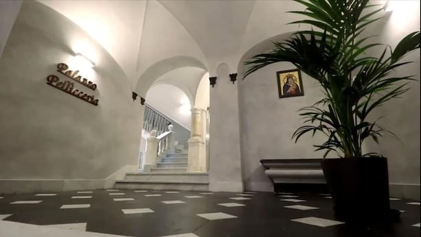 Um corredor com piso em padrão preto e branco, uma grande planta em vaso e uma escadaria que leva a um andar superior.