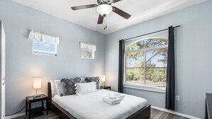 Quarto com cama, duas mesas de cabeceira, ventilador de teto e uma janela grande com cortinas.
