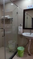 Casa de banho com cabine de chuveiro em vidro, pia, um cesto de lixo verde e um porta-toalhas.