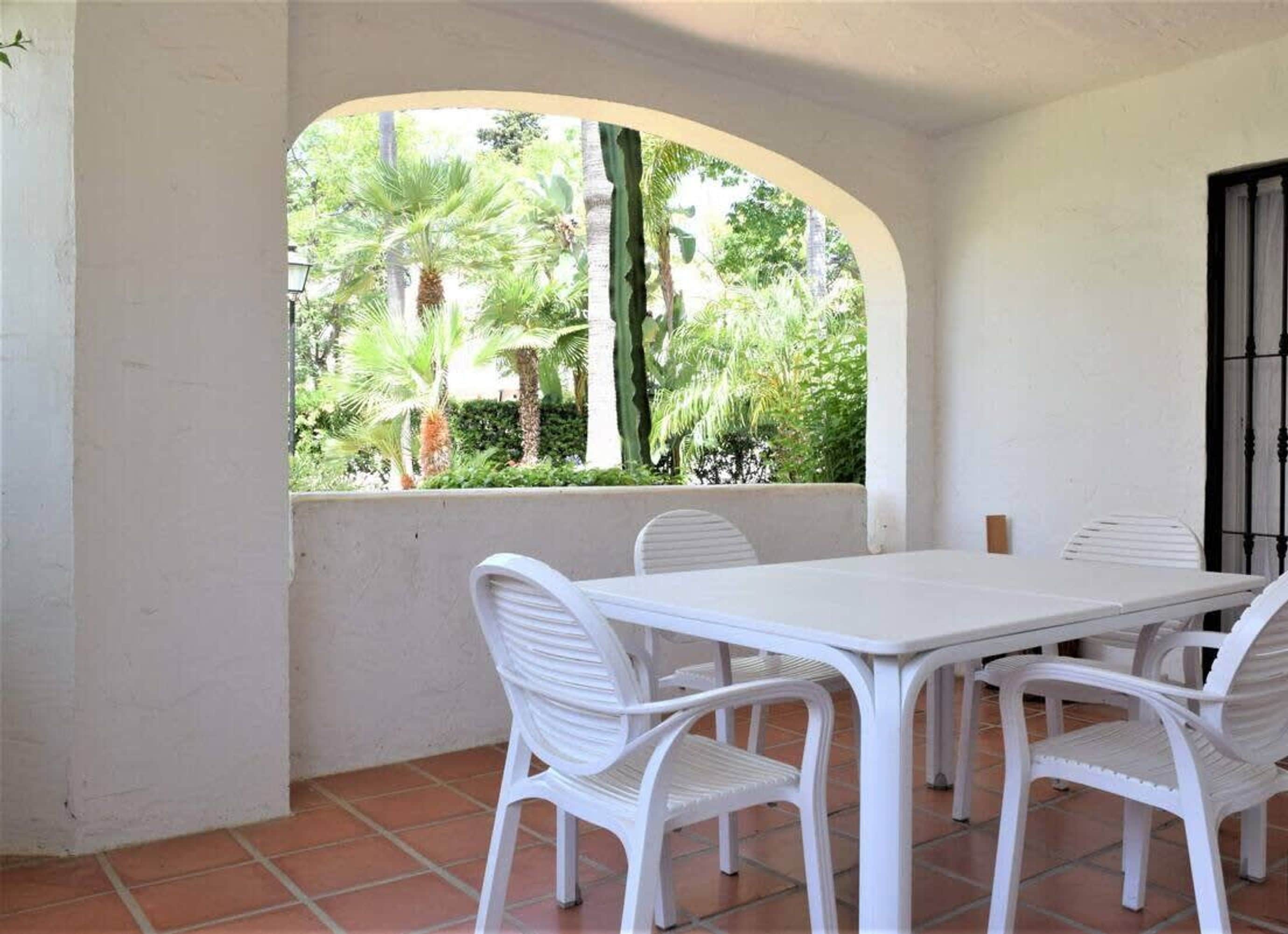 Um pátio branco com mesa e cadeiras, vista para o verde através de uma janela em arco e um portão preto.