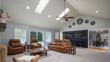 Uma sala de estar espaçosa com teto alto, ventilador de teto e janelas grandes. Há um quadro negro com escrita a giz na parede e uma área de jantar com mesa e cadeiras.