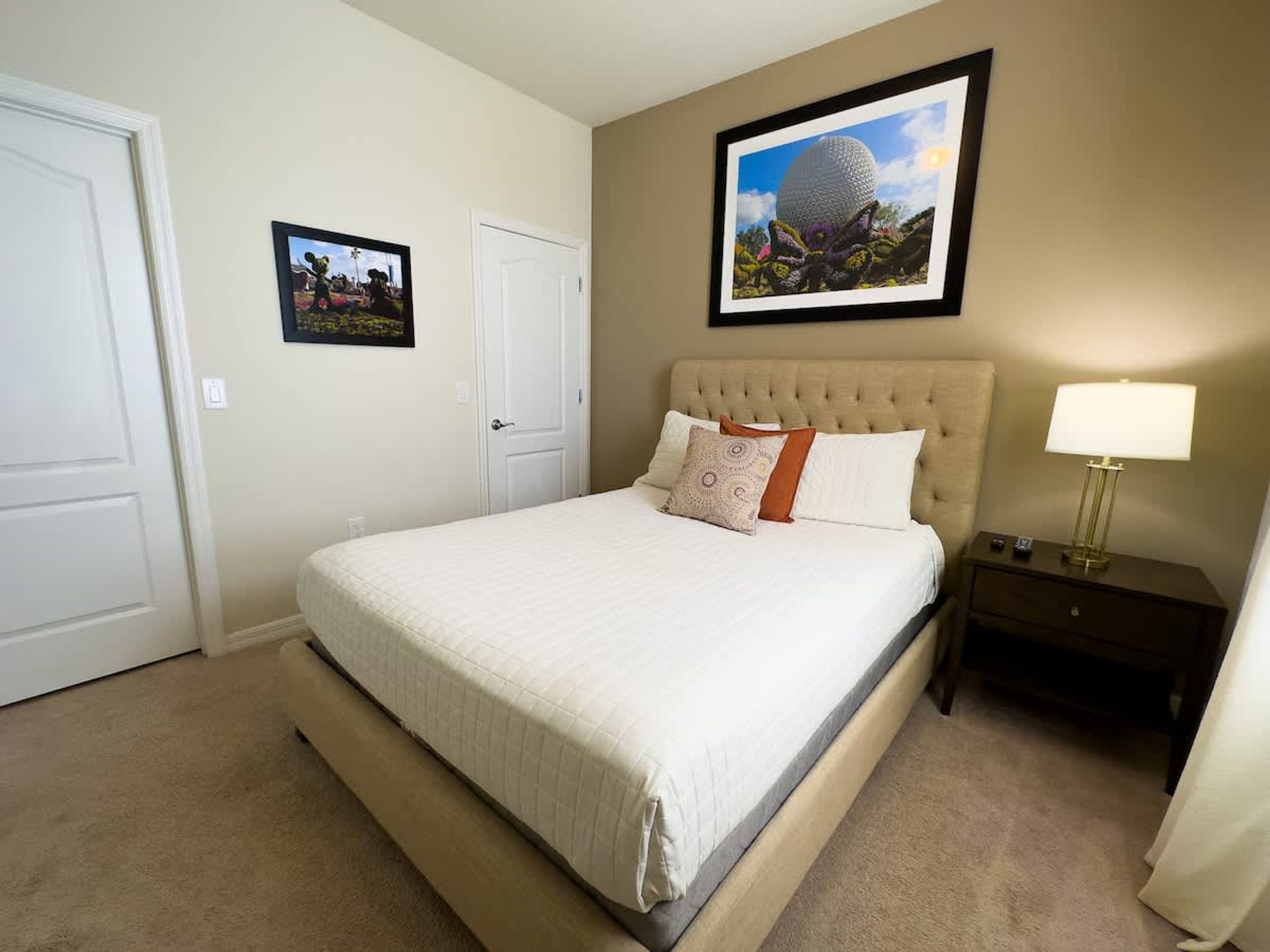 Quarto de hotel com uma cama grande, uma mesa de cabeceira com abajur e duas fotos emolduradas na parede.