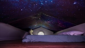 Quarto com teto inclinado, mural com tema de galáxia, janela e cama com almofadas.