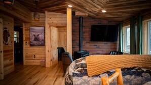 Interior de uma cabana de madeira com uma cama, lareira, TV na parede e teto de madeira.