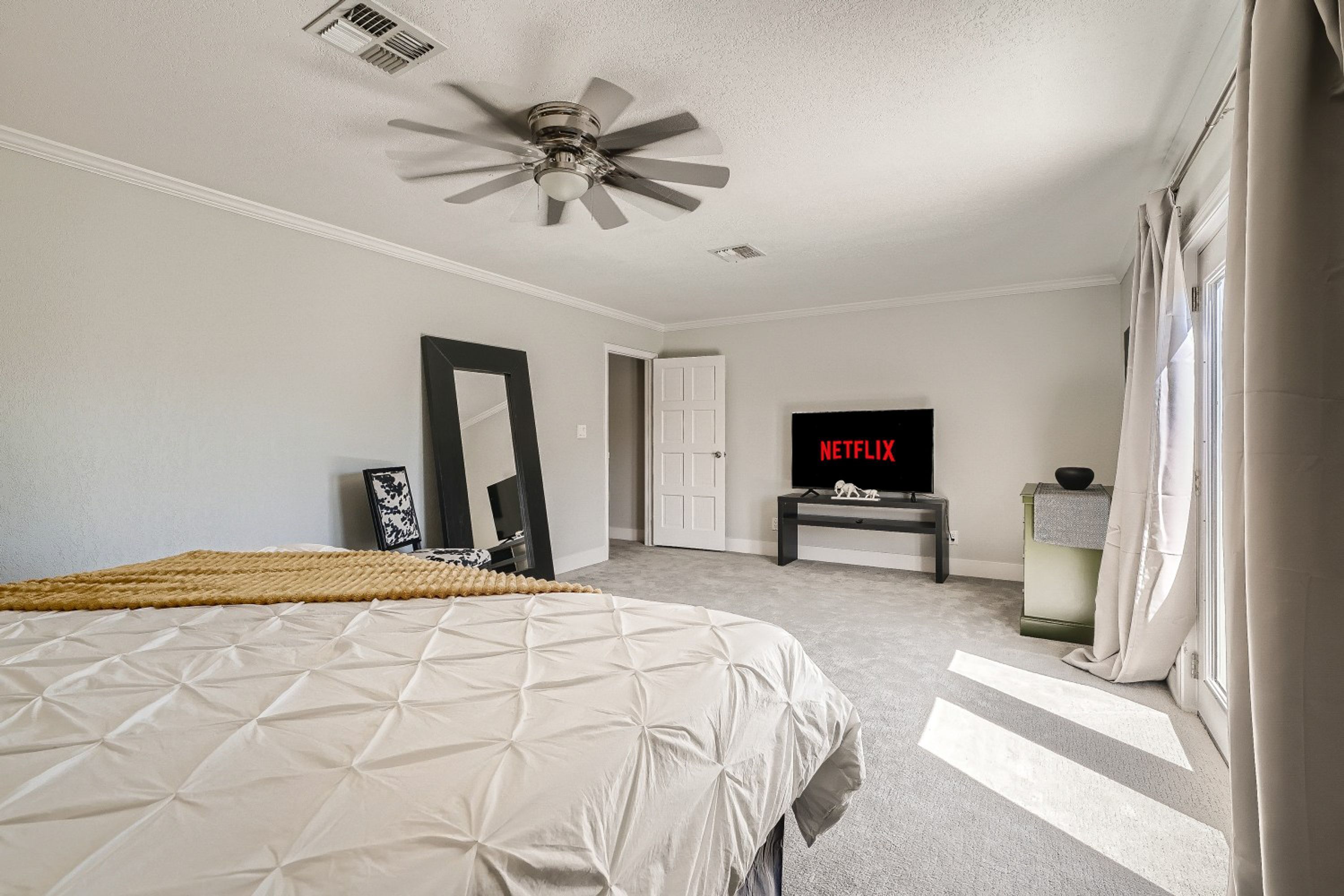 Quarto com cama, ventilador de teto, TV exibindo 'NETFLIX' e porta que dá para outro cômodo.