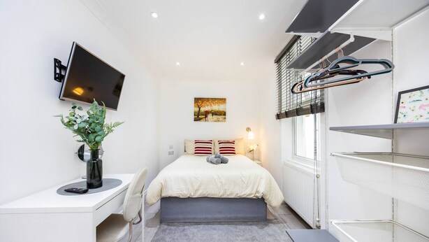 Quarto compacto com cama, mesa de cabeceira, televisão e pequena área de trabalho.